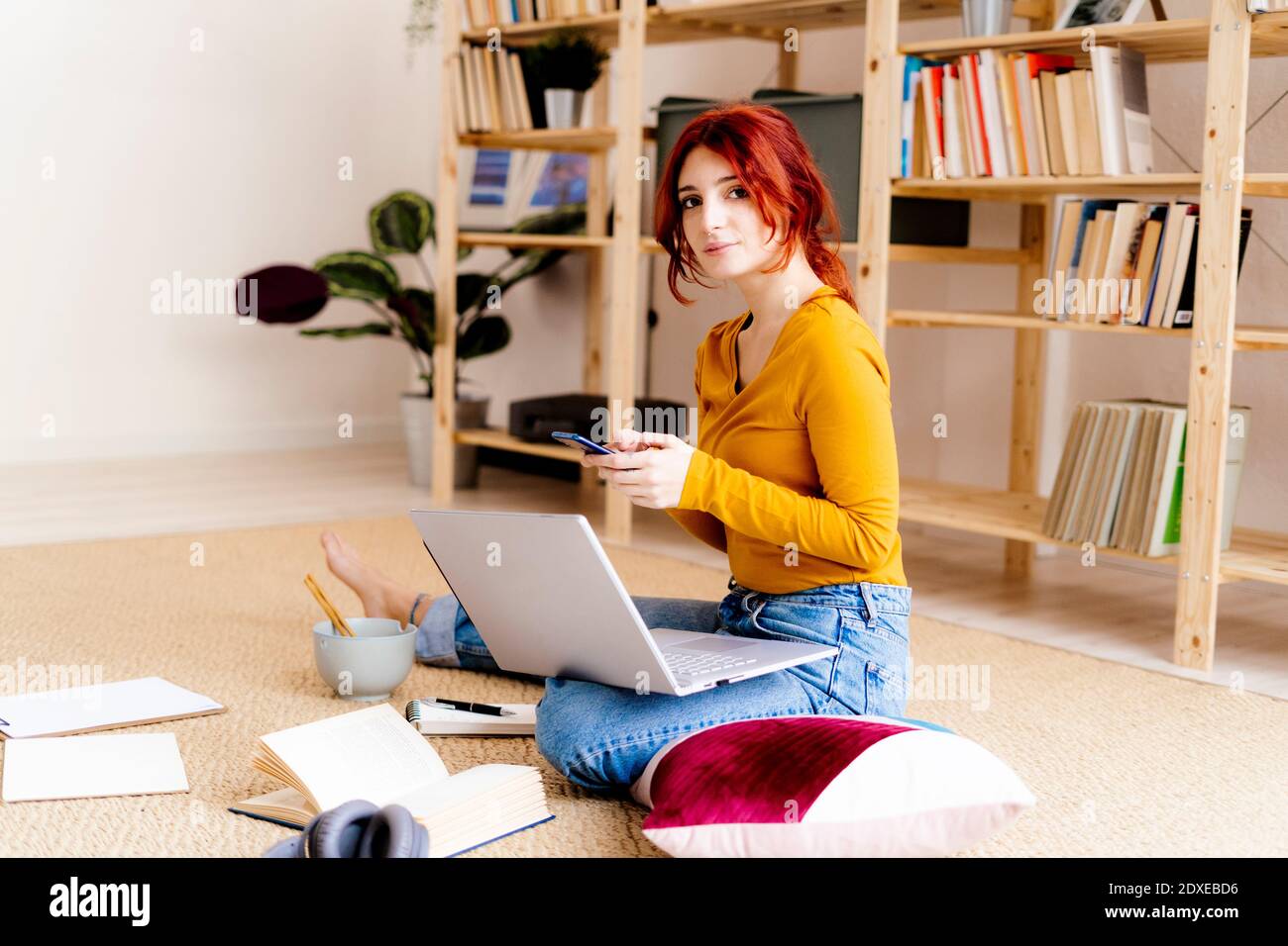 Young woman with laptop on lap using mobile phone while sitting at home ...
