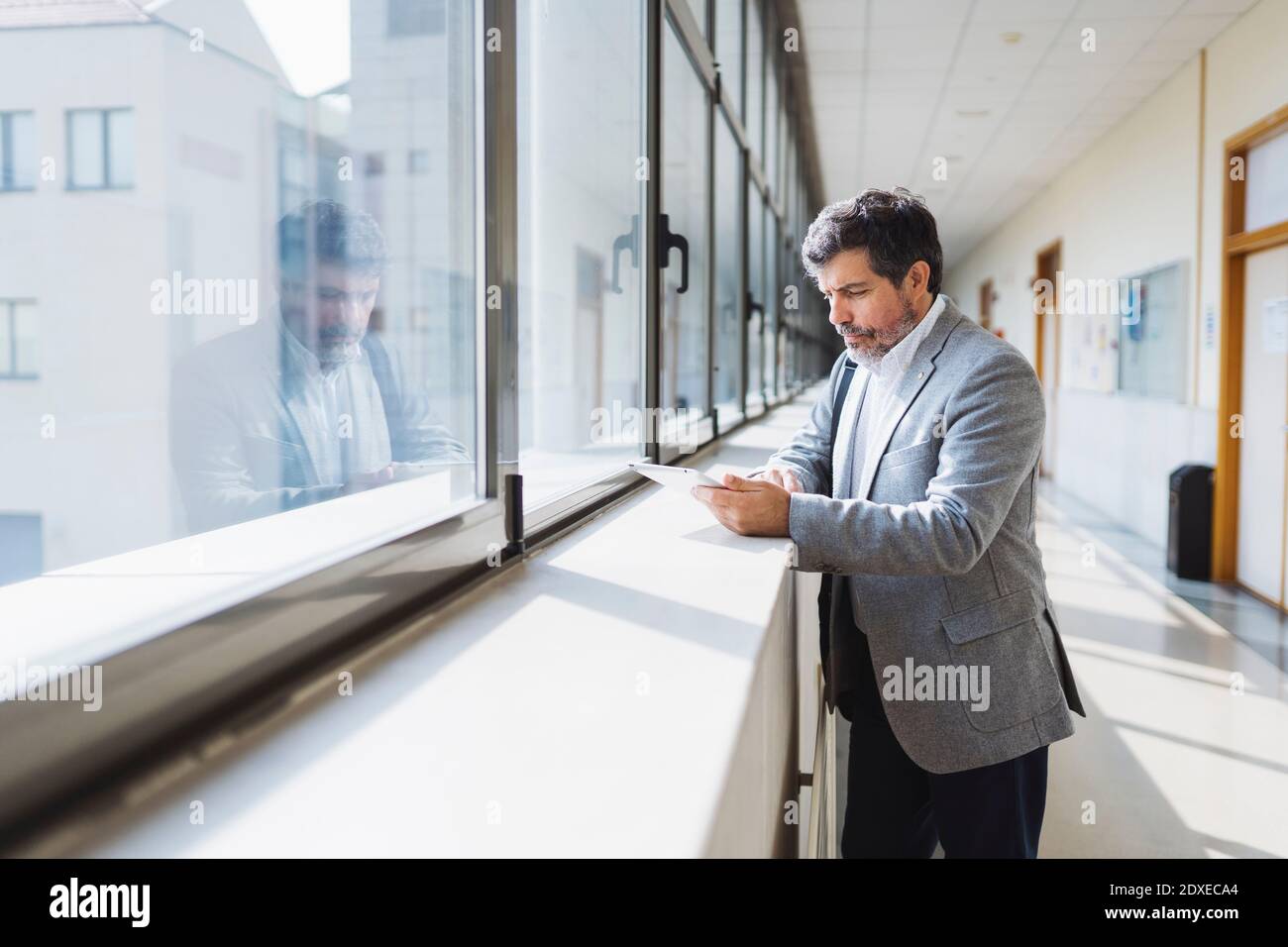 Professor using digital tablet standing by window sill in corridor at ...