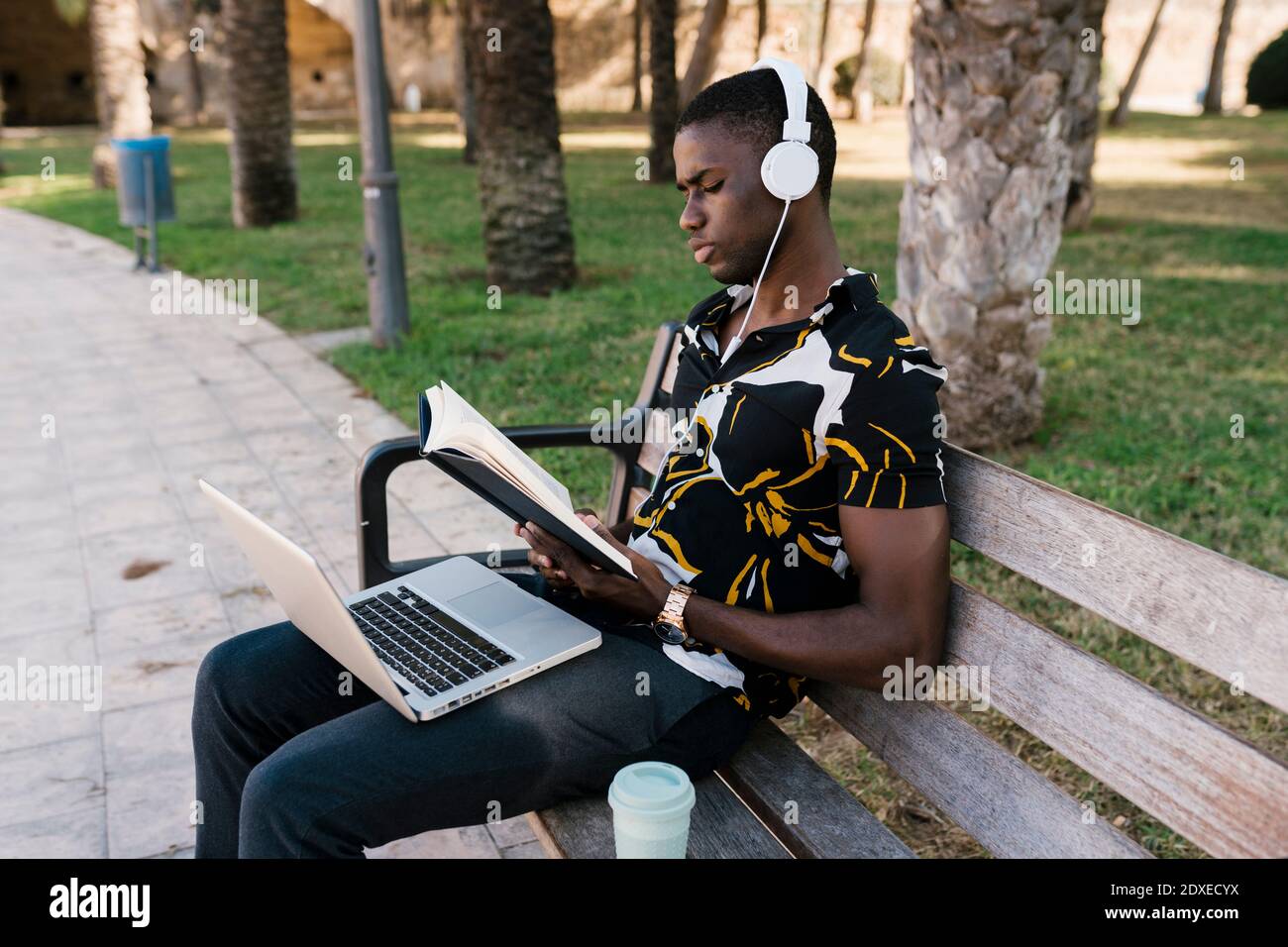 Businessman with laptop reading book while sitting on bench in park ...