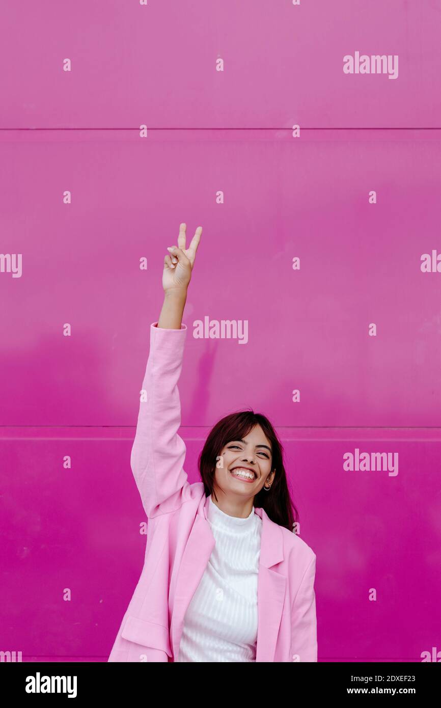 Smiling woman with hand raised doing peace sign while standing against ...
