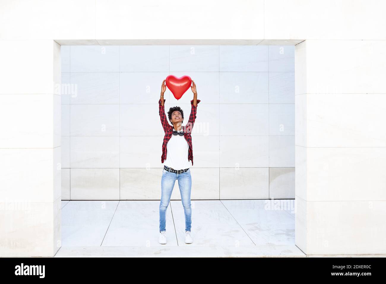 Young woman holding aloft red heart shape balloon against white wall ...