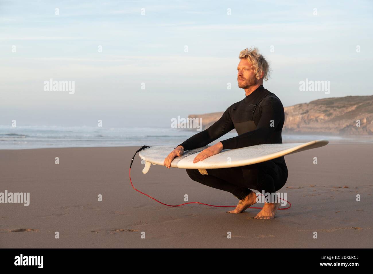 Blond surfer with surfboard on lap crouching at beach against sky Stock ...