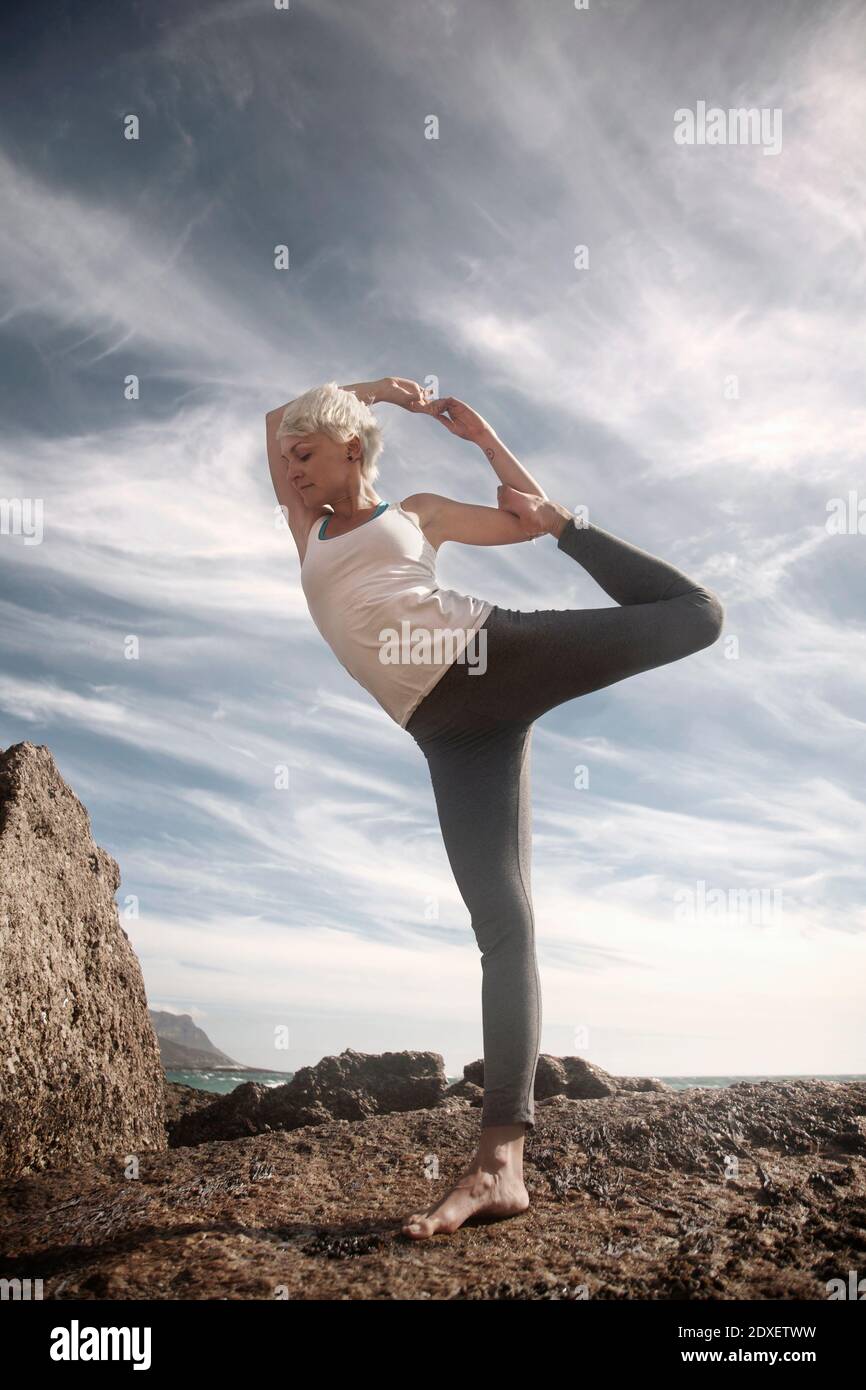 Flexible woman practicing dancer's pose yoga on rock formation at beach ...