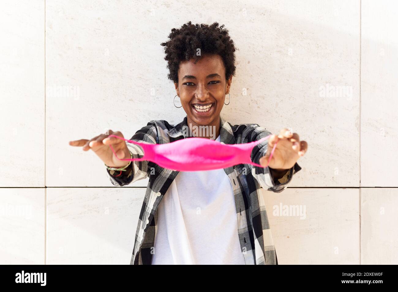 Cheerful young woman pulling pink mask against wall Stock Photo - Alamy