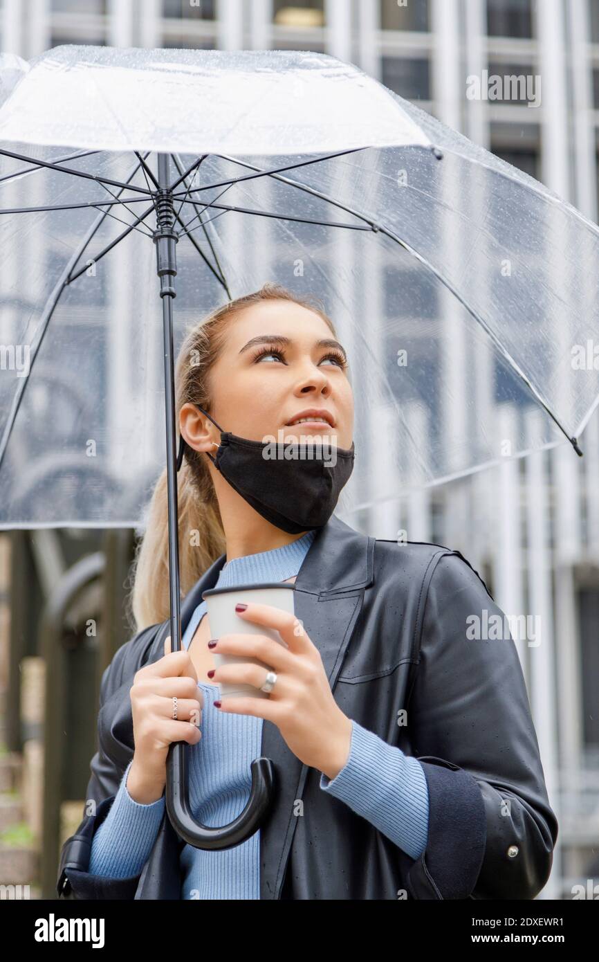 Thoughtful businesswoman wearing face mask holding umbrella and ...