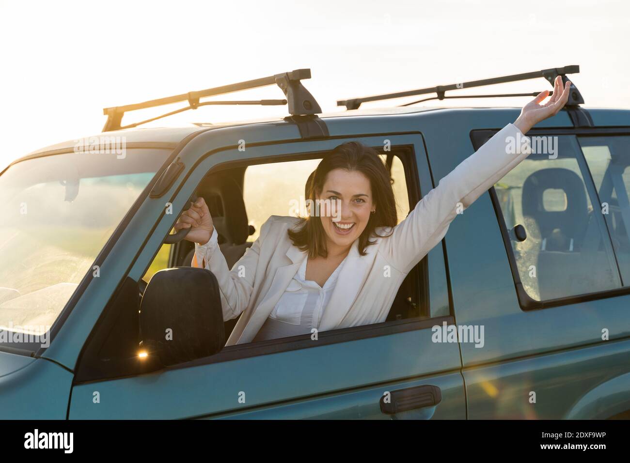Carefree young woman leaning out of car window with arms raised at ...