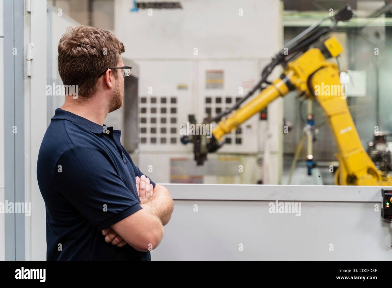 Male factory worker with arms crossed looking at robotic arm through ...