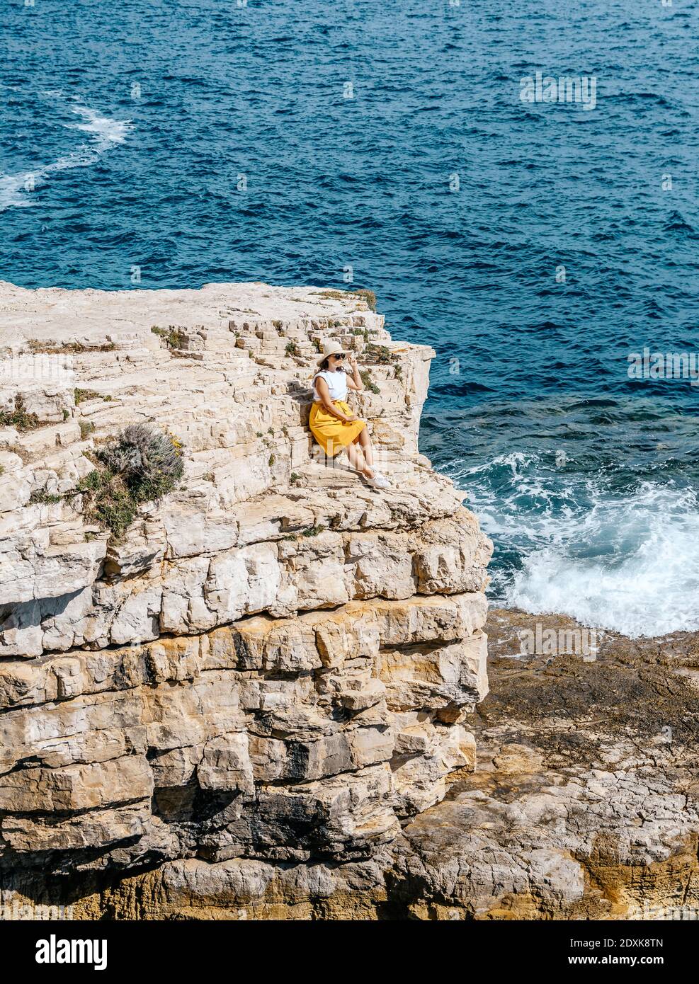 A vertical shot of a female sitting on the cliff in the coastal area in ...