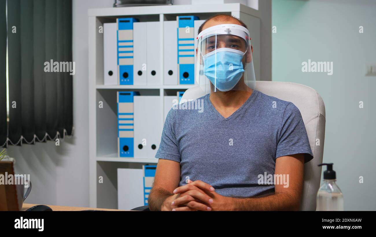 Busy employee wearing mask and visor in work place, typing and looking ...