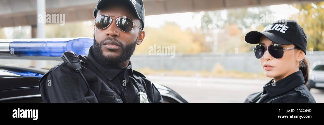 multicultural police officers looking away near patrol car on blurred ...