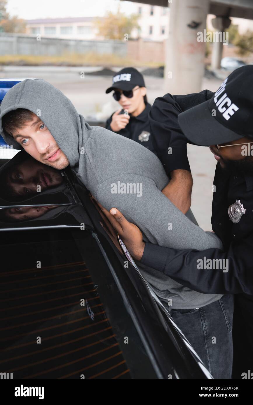 african american policeman frisking sad hooded offender leaning on car ...