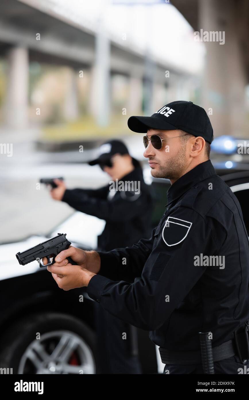 Police officer in sunglasses holding firearm near colleague and car on ...