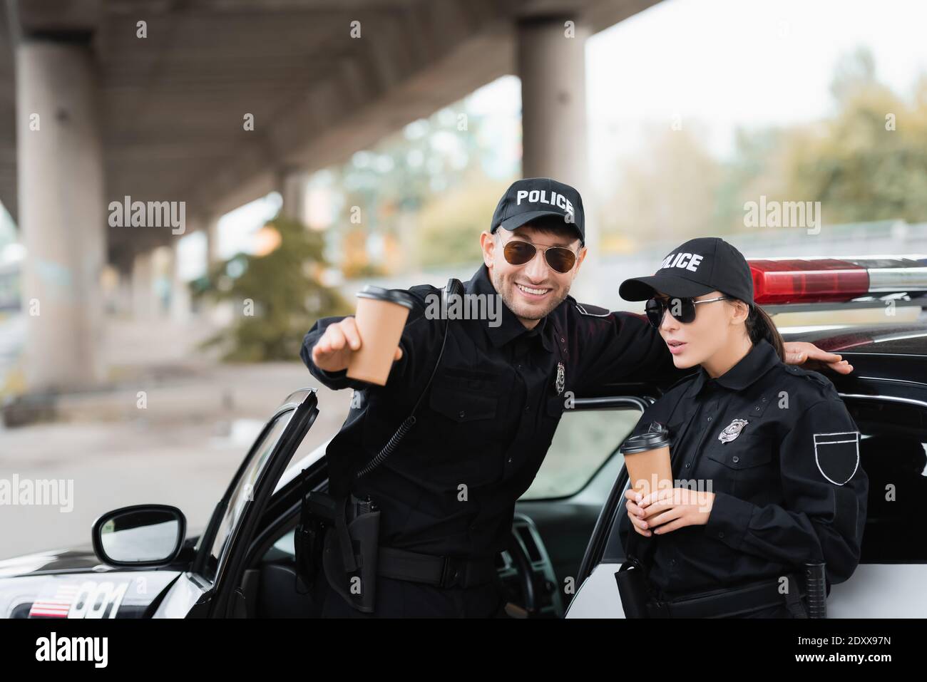 Cheerful police officer pointing with hand near colleague holding ...