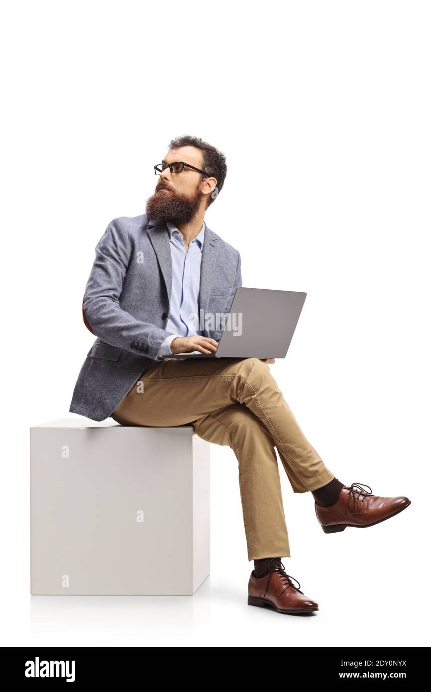 Bearded man sitting on a white cube with a laptop computer and looking ...