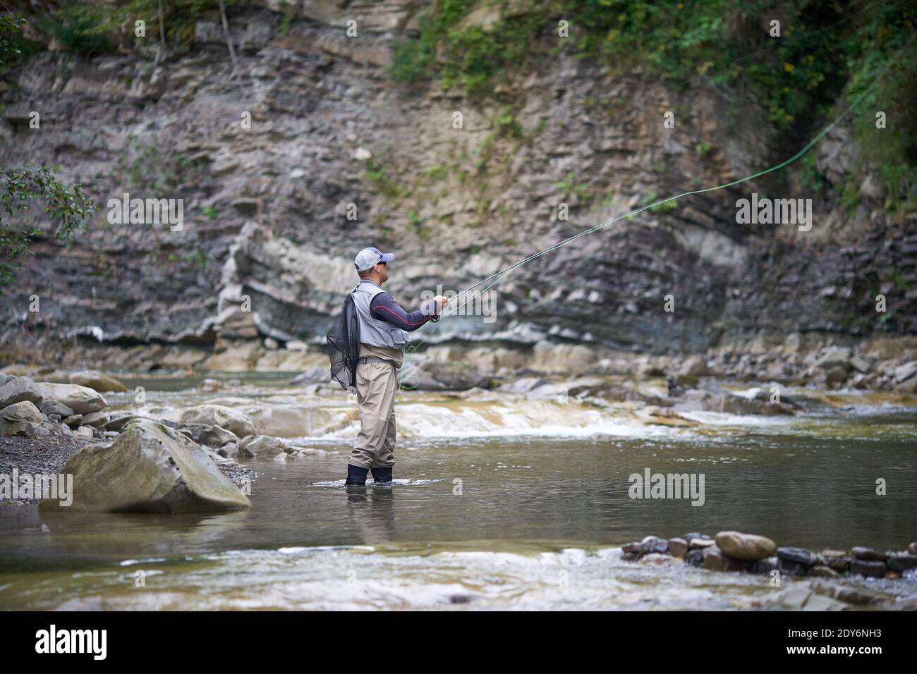 Competent fisherman in uniform and cap hooking with rod in rough river ...