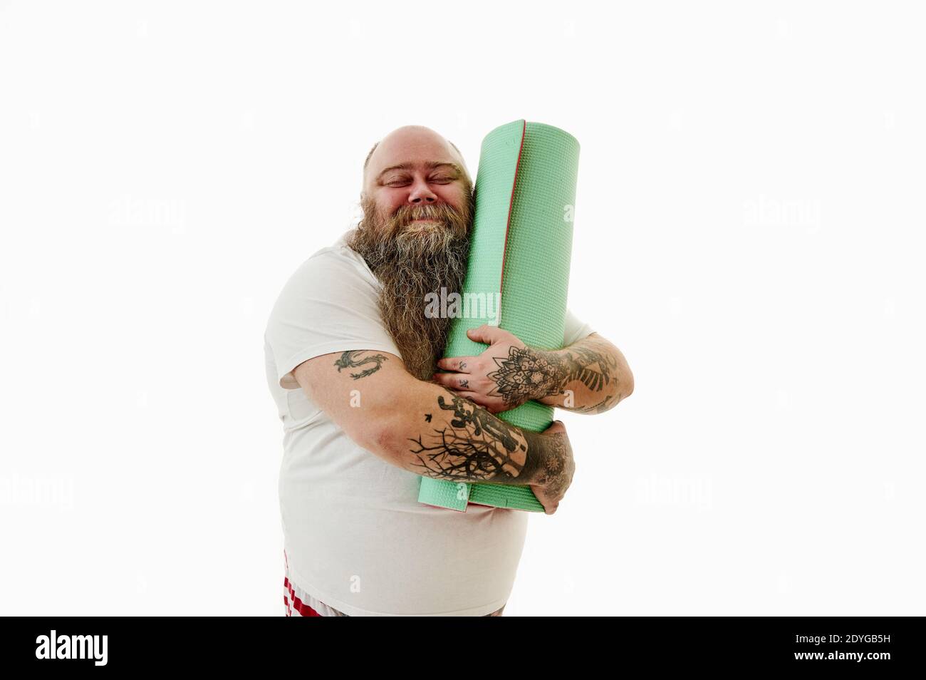 A happy overweight man with tattooed arms gently hugs his fitness mat ...