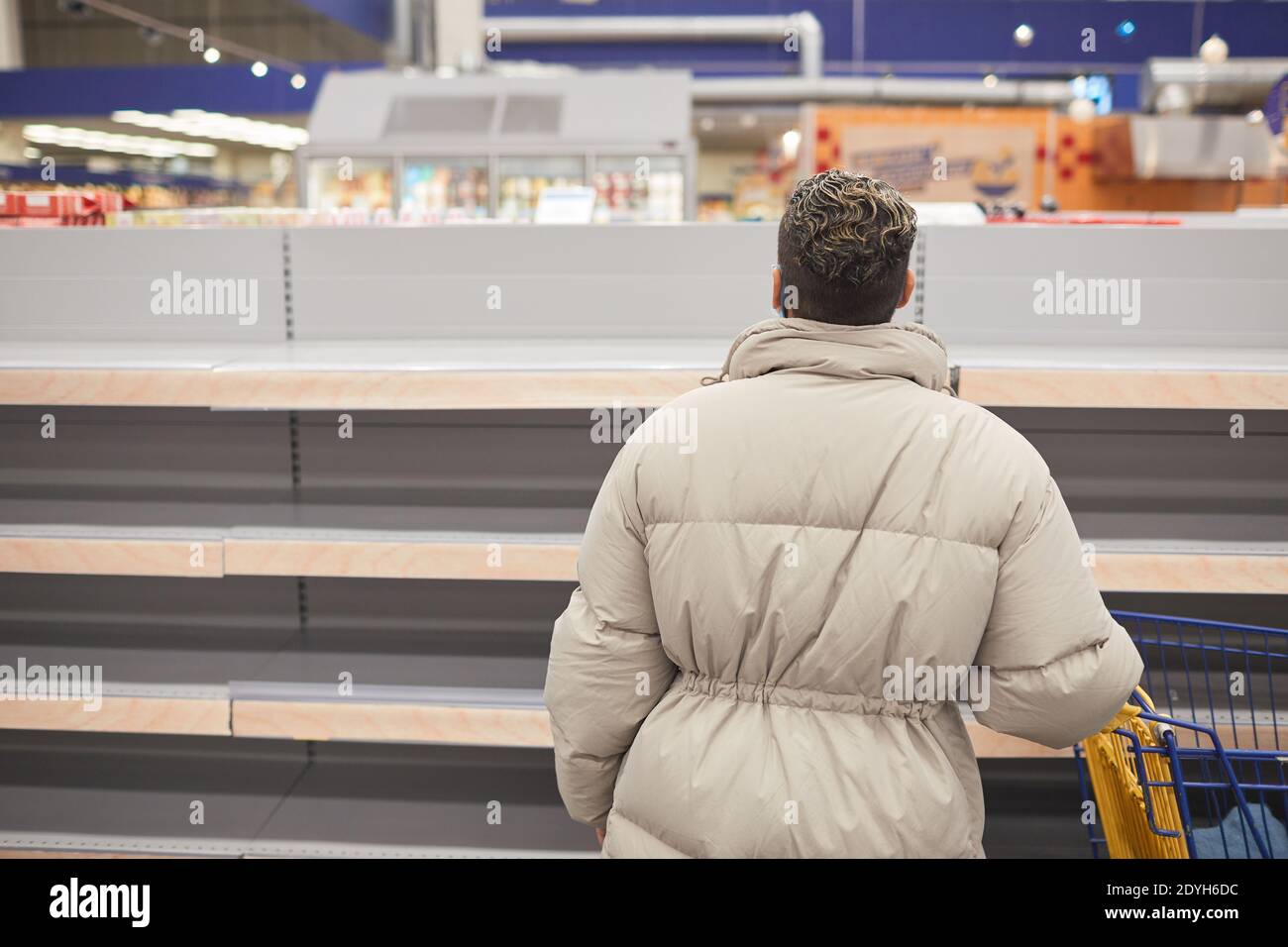 Rear view of customer with shopping cart looking at empty shelves while ...