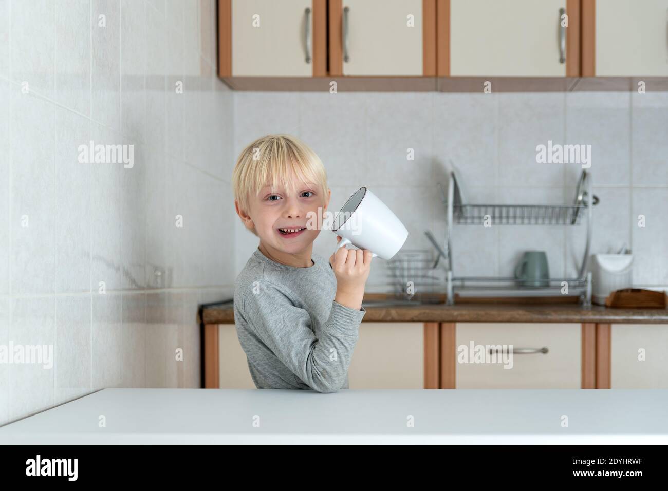 Blond boy drinking from large cup in kitchen. Funny schoolboy breakfast ...