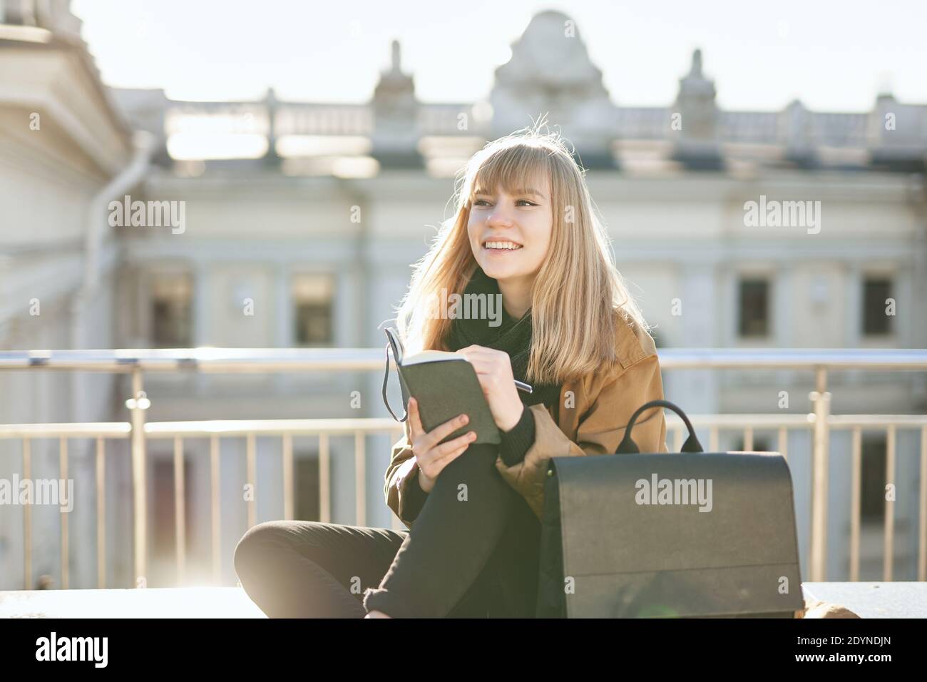 Beautiful blonde young girl in a trench sitting on the roof and writing ...