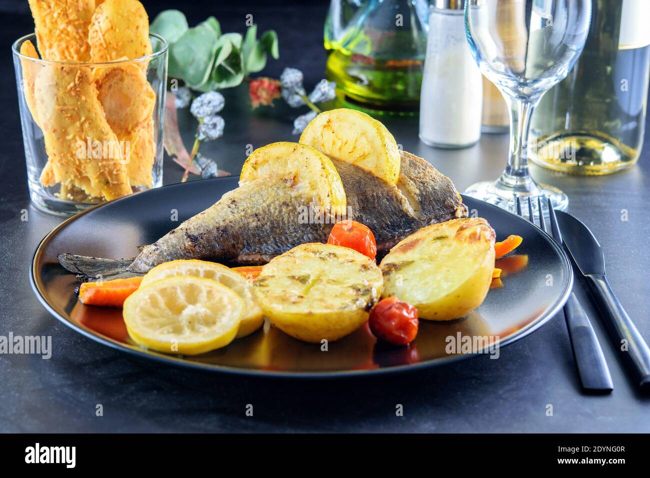Black dish with fried fish dorada, slices of lemon, potatoes, carrot ...