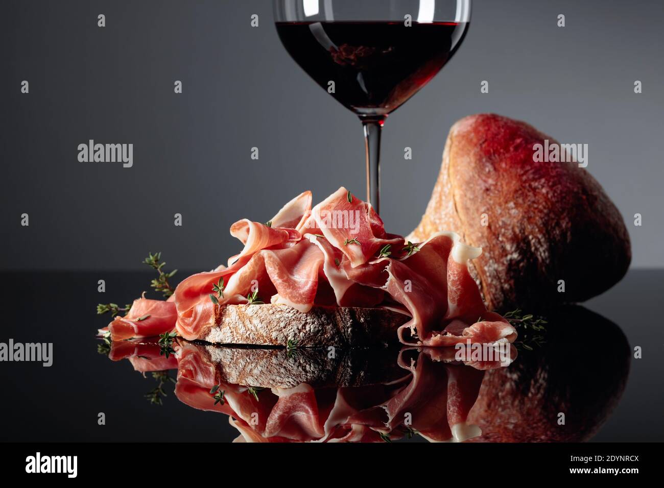 Prosciutto with ciabatta, red wine, and thyme on a black reflective ...
