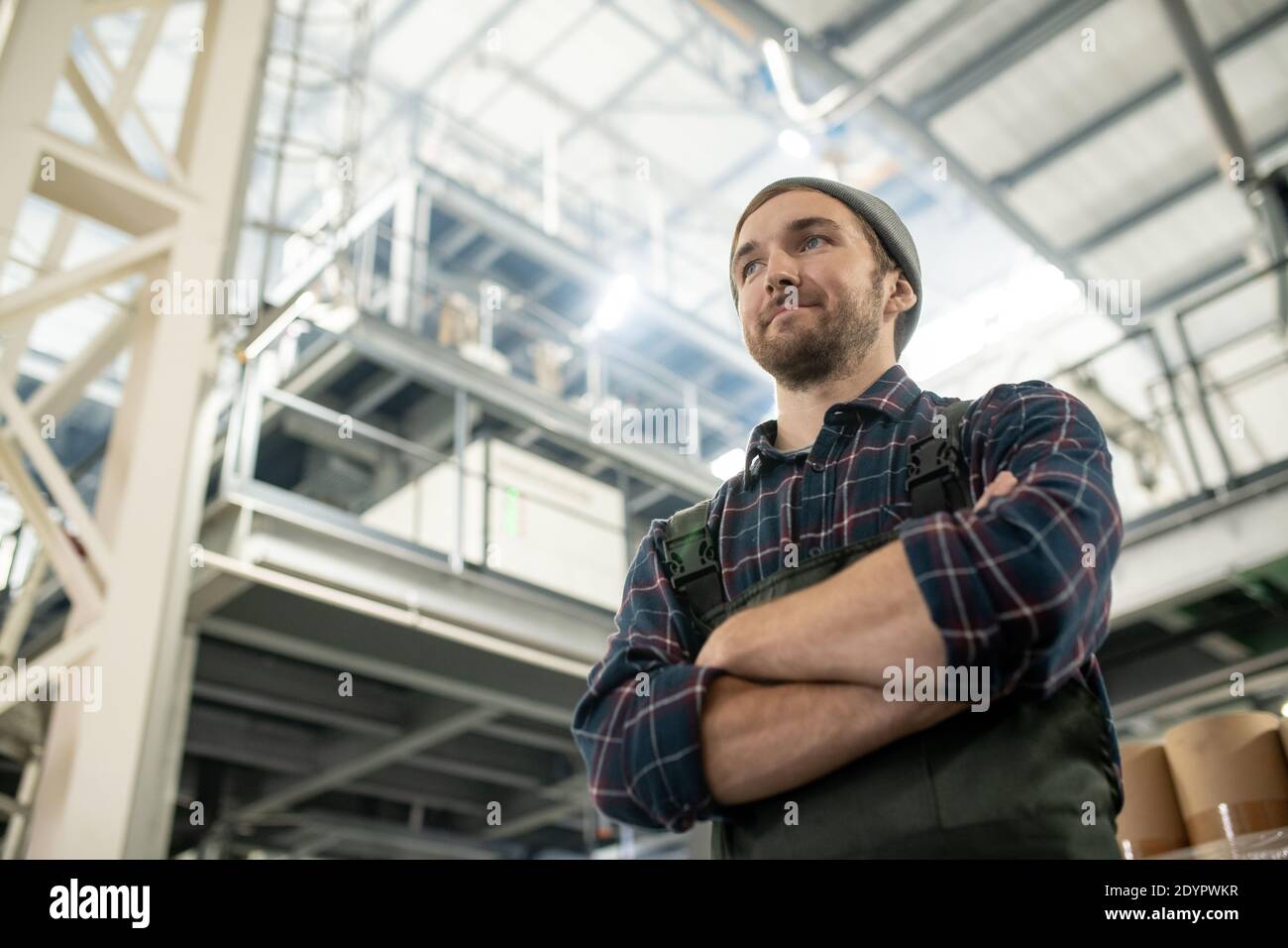 Young confident male worker of large factory in shirt, beanie and ...