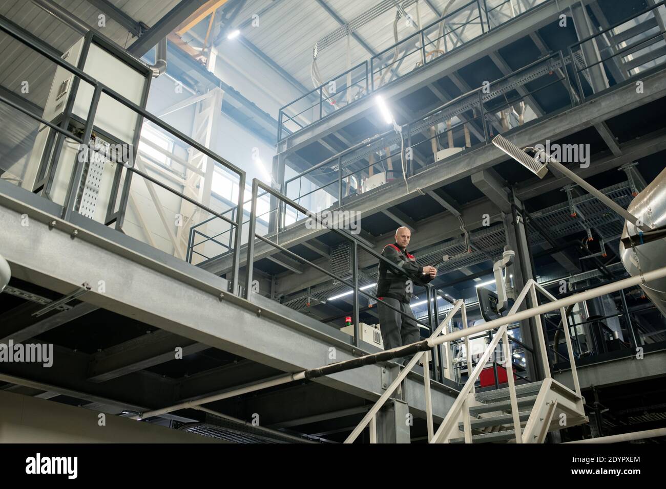 Bald mature engineer or foreman of contemporary industrial plant standing inside large polymer ...