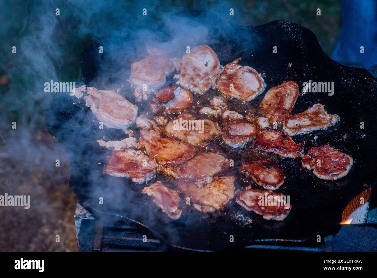 Fresh cut bacon being cooked over a campfire. The meat is fried in a ...