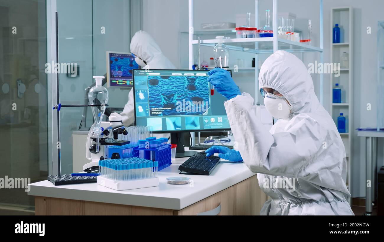 Chemist in coverall checking blood samples taking notes on computer in ...