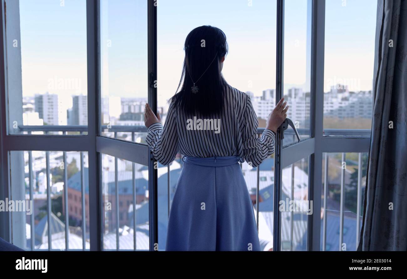 Back view of young woman looking out window, sunny weather and city ...