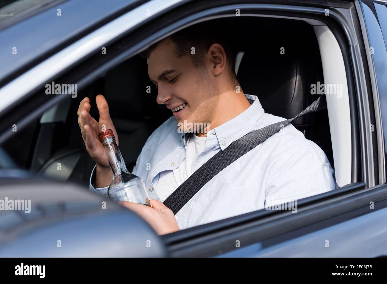 drunk man smiling and holding bottle of whiskey while sitting in car on ...