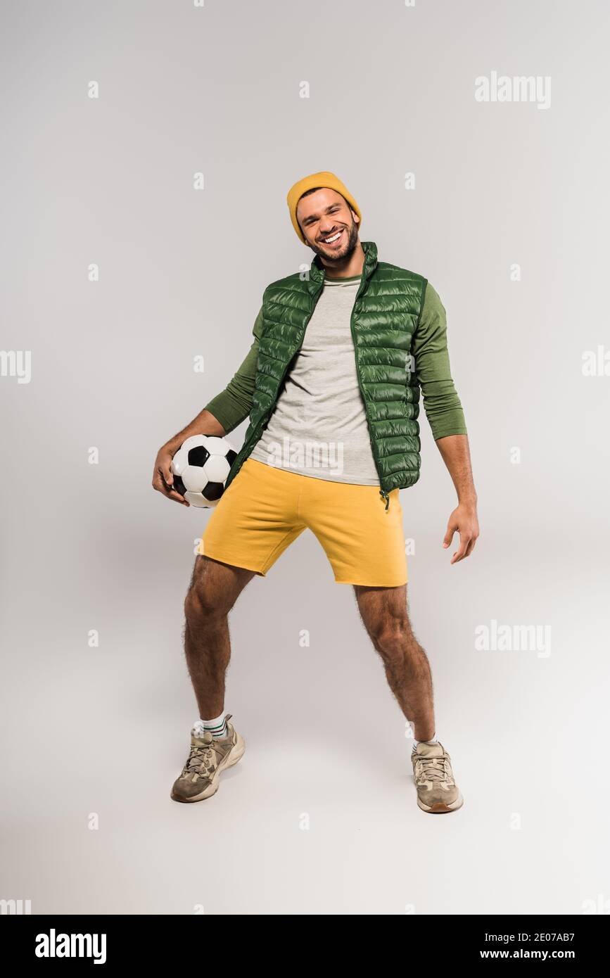 Positive sportsman in casual clothes holding football on grey ...