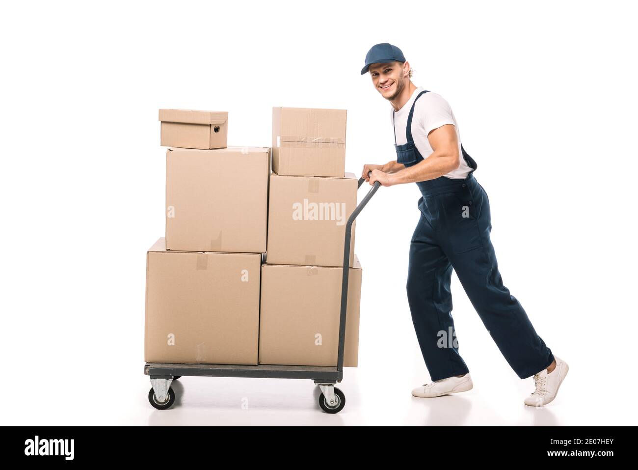 full length of cheerful mover in uniform and cap pulling hand truck ...