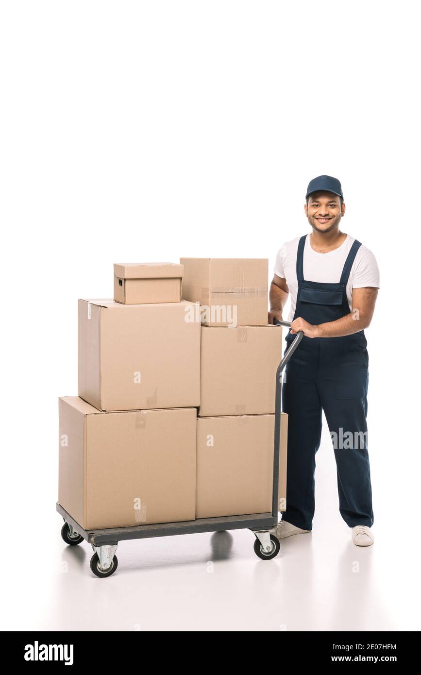 full length of happy indian mover in overalls standing near hand truck ...