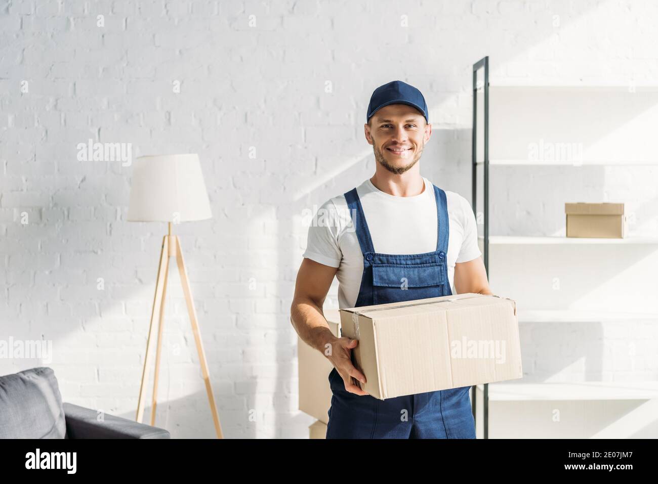 cheerful mover in cap holding carton box in apartment Stock Photo - Alamy