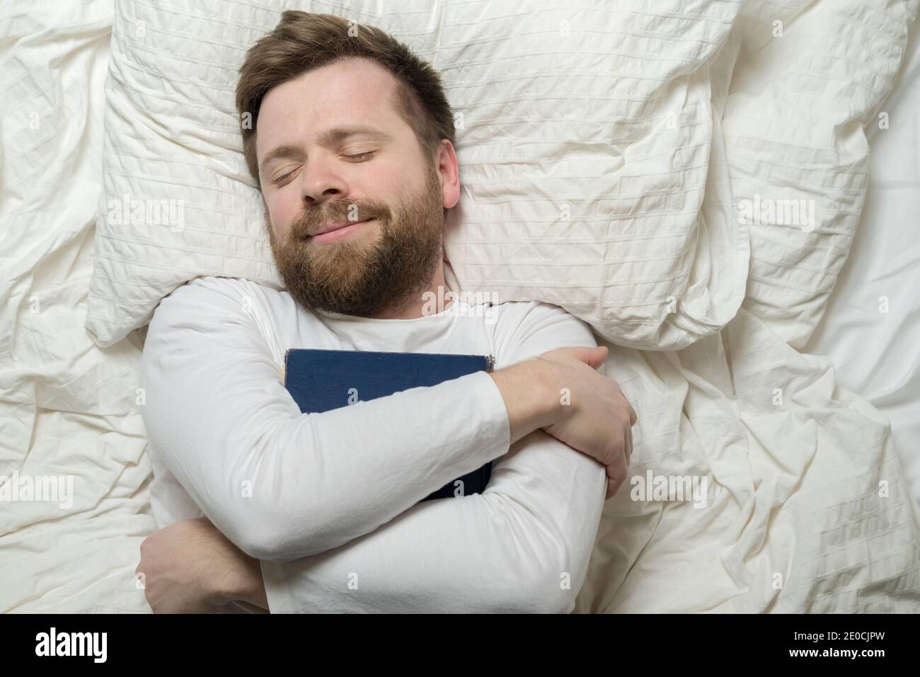 Charming man hugs a book while lying in bed. He dreams with his eyes ...