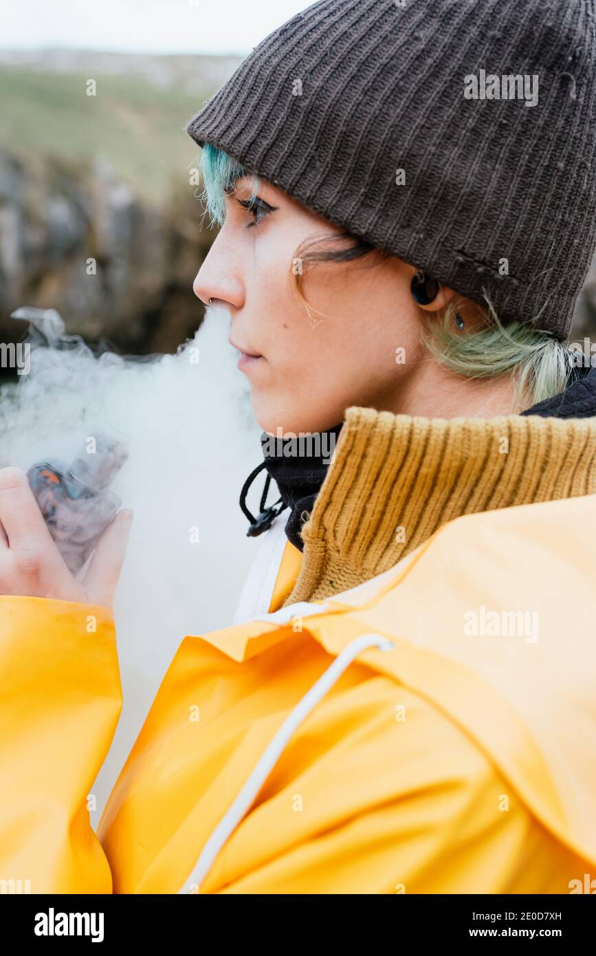 Side view of relaxed female in outerwear smoking vape and exhaling ...