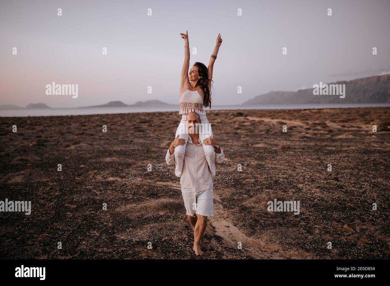 Cheerful Woman with raised arms sitting on shoulders of smiling man ...