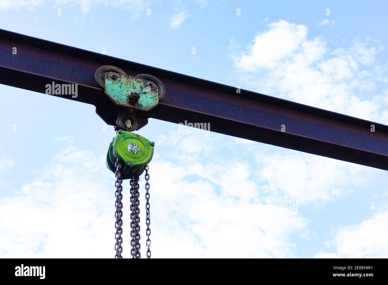 Green chain fall hoist on an I-beam silhouetted against a pale blue sky ...