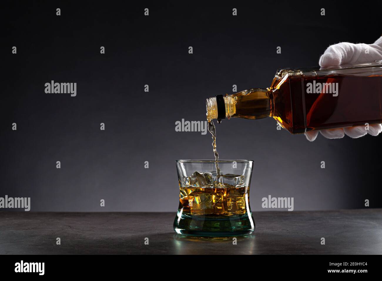 White gloved hand pouring whisky into a glass with ice on dark ...