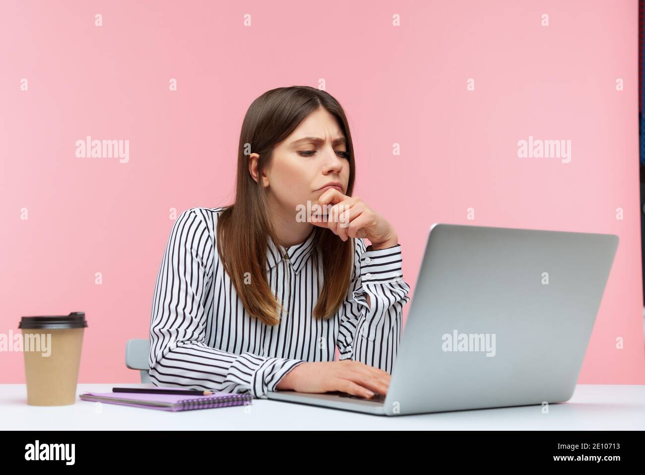 Puzzled brunette business woman in striped shirt sitting at workplace ...