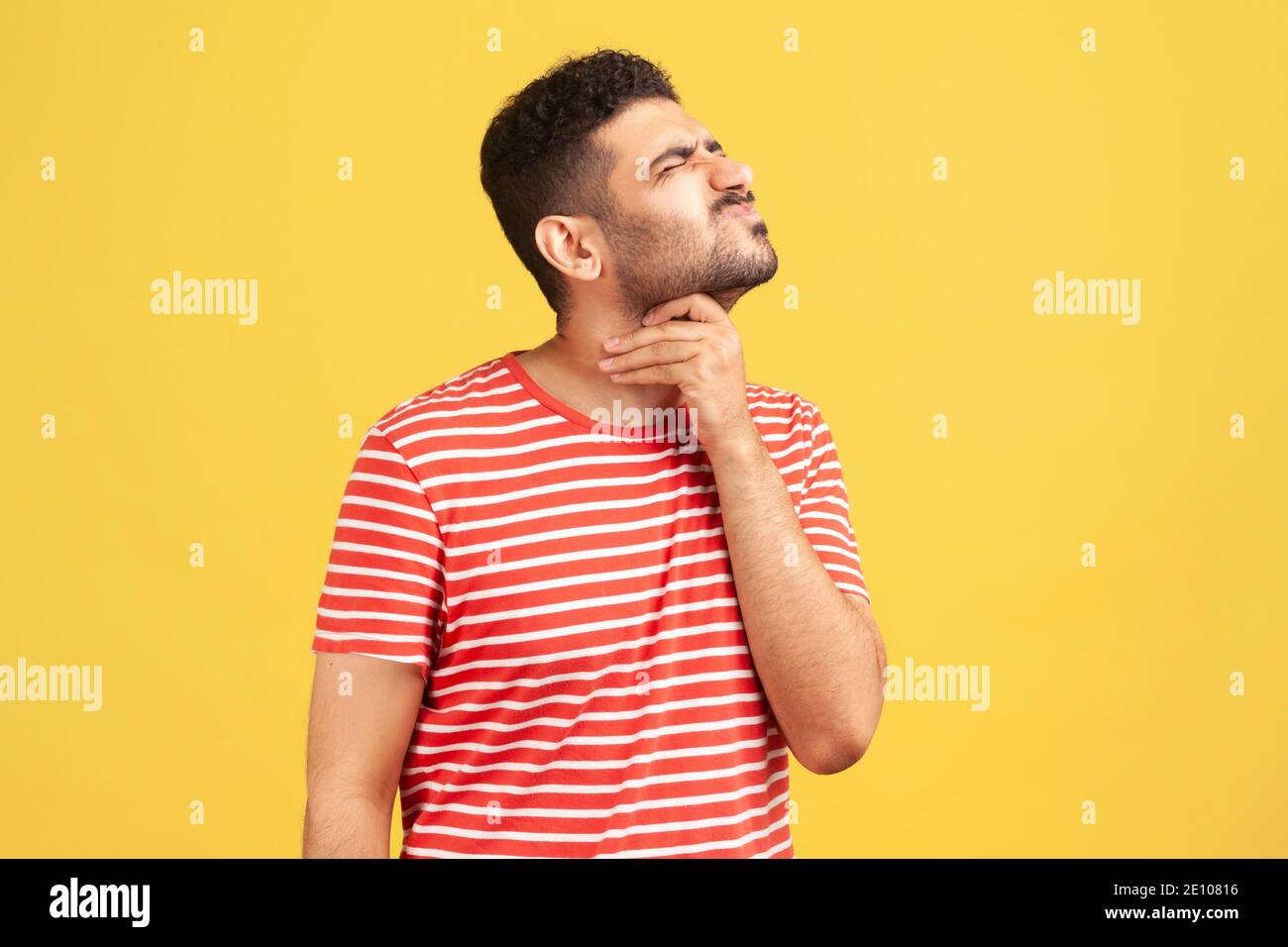 Unhappy bearded man in striped t-shirt grimacing touching his neck ...