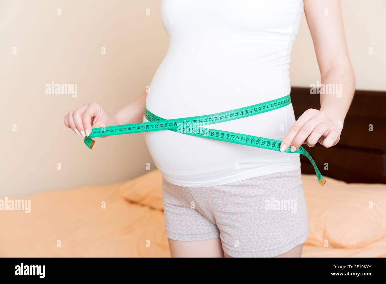 Young beautiful pregnant girl measuring her belly with a tape in home ...