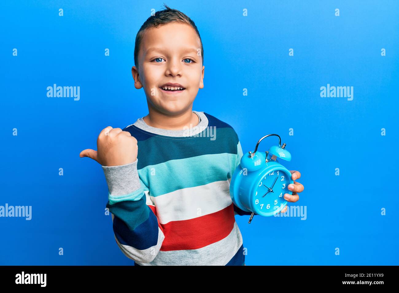 Little kid boy holding alarm clock pointing thumb up to the side ...