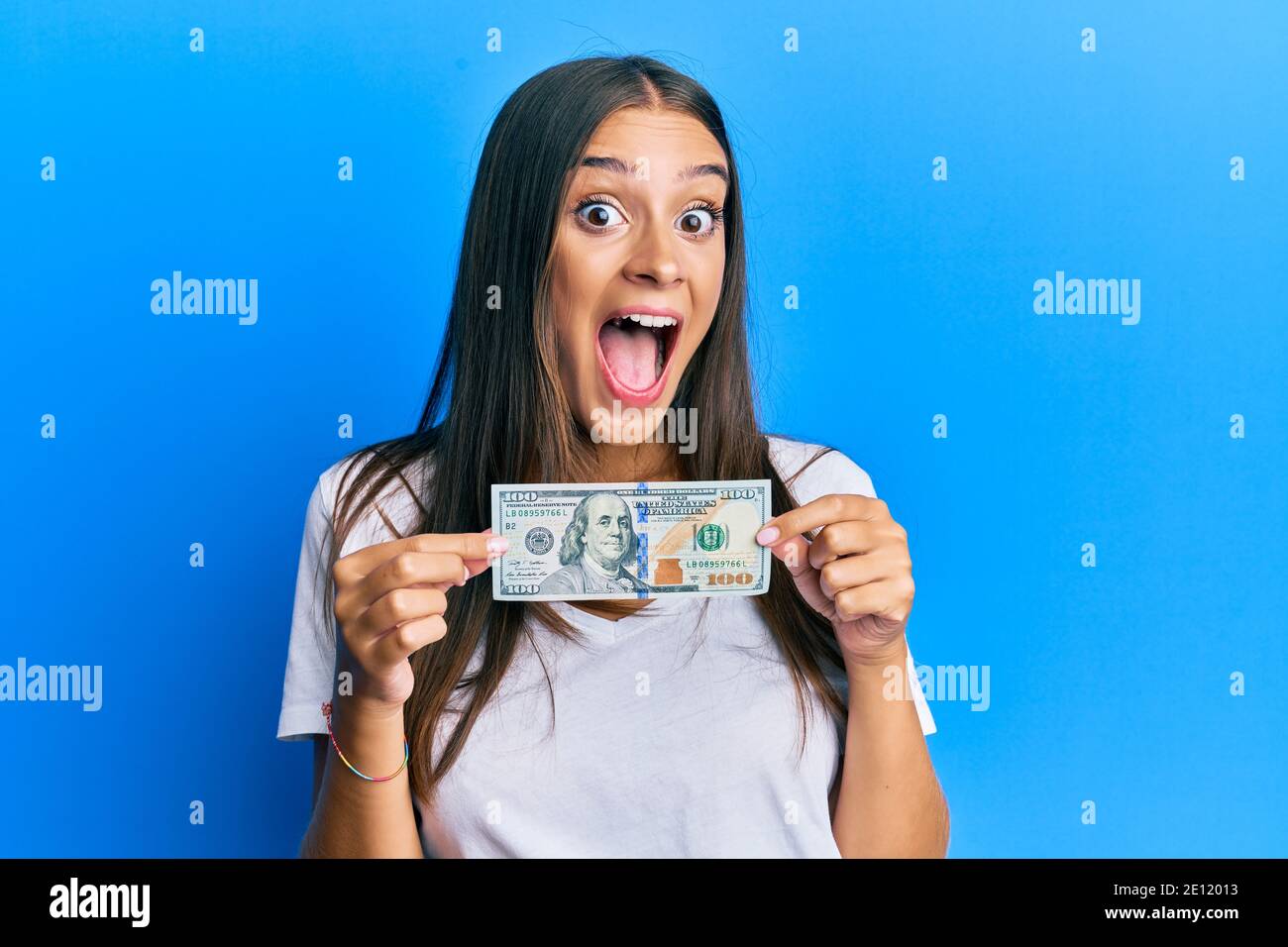 Young hispanic woman holding 100 dollar banknote celebrating crazy and ...