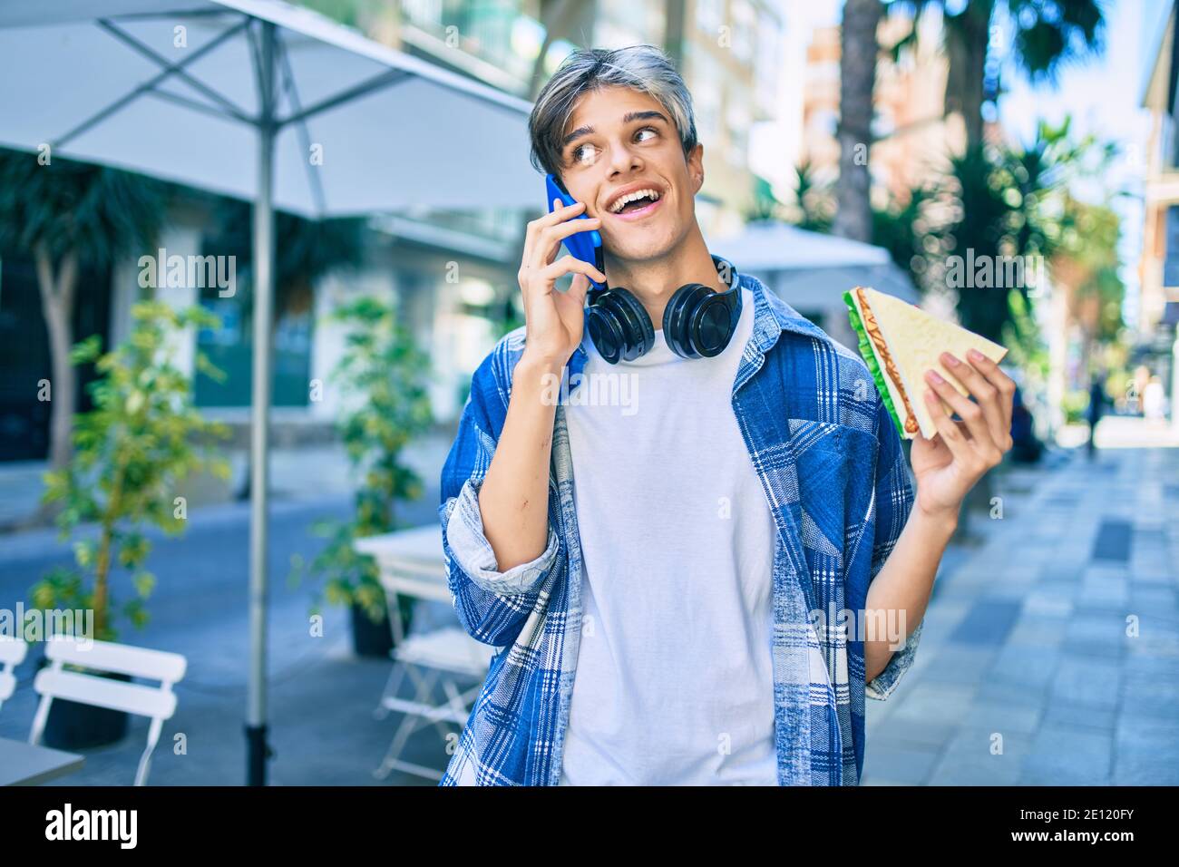 Young hispanic man smiling happy talking on the smartphone and eating ...