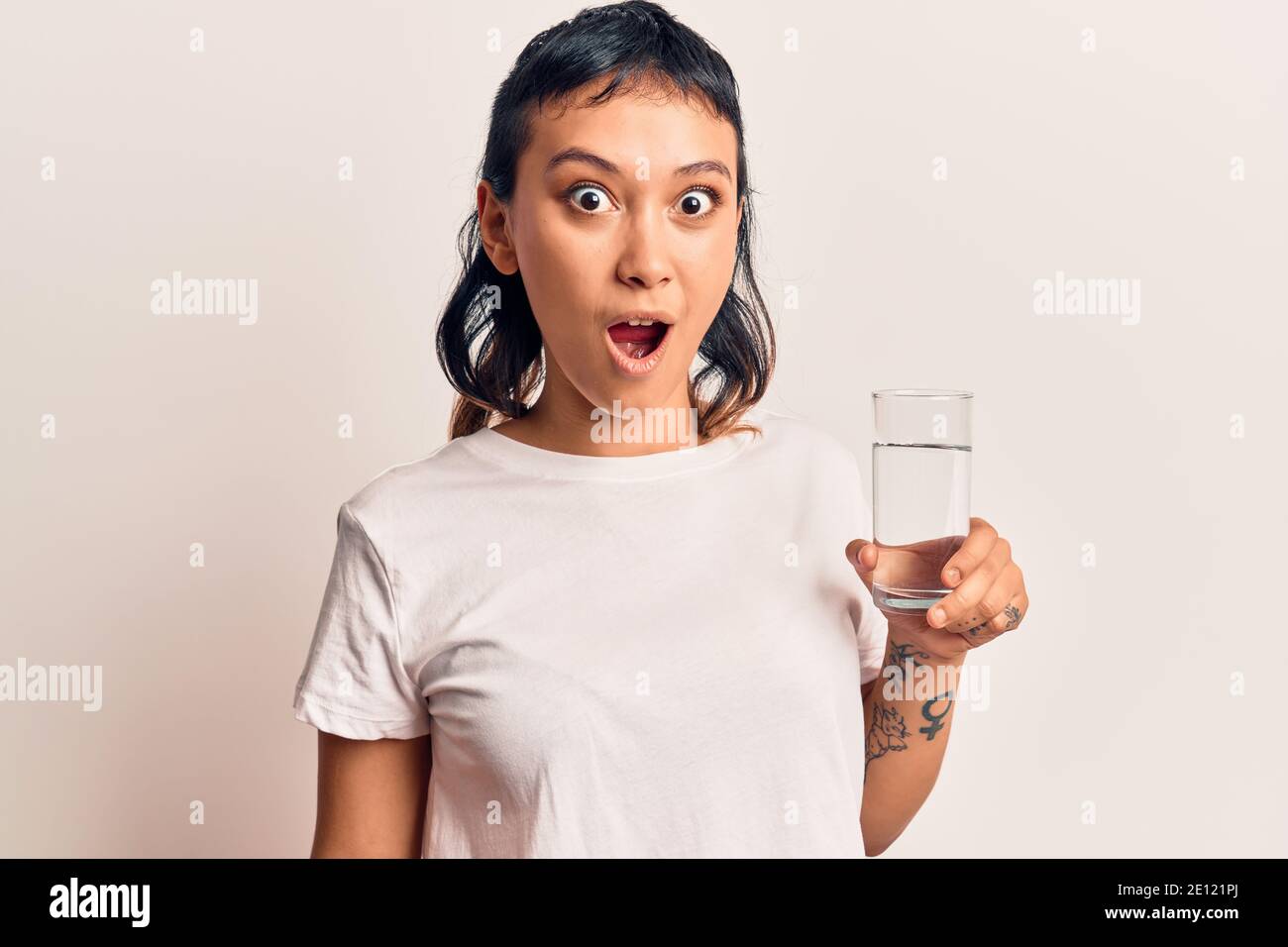 Young woman drinking glass of water scared and amazed with open mouth ...