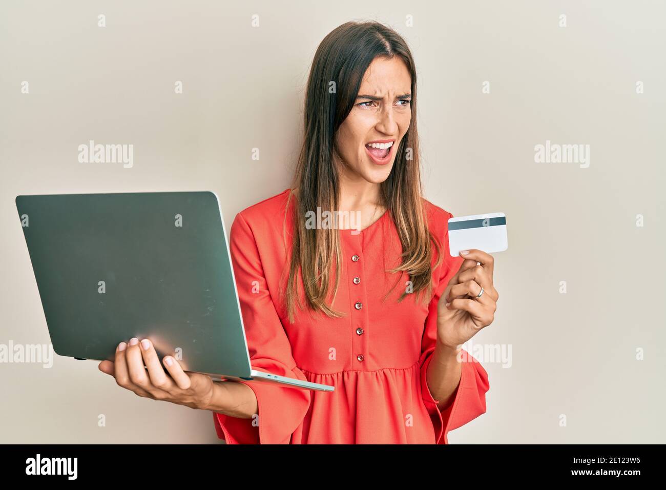 Young beautiful woman holding laptop and credit card angry and mad ...