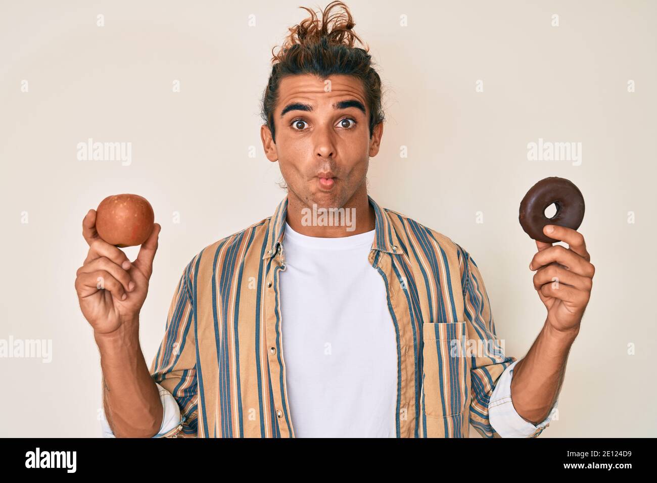 Young hispanic man holding red apple and donut making fish face with ...