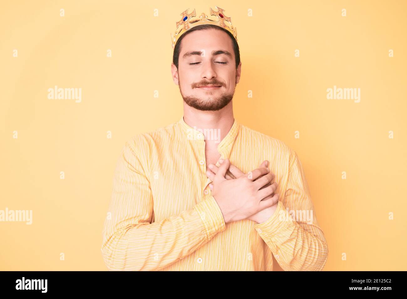 Young handsome man wearing king crown smiling with hands on chest, eyes ...